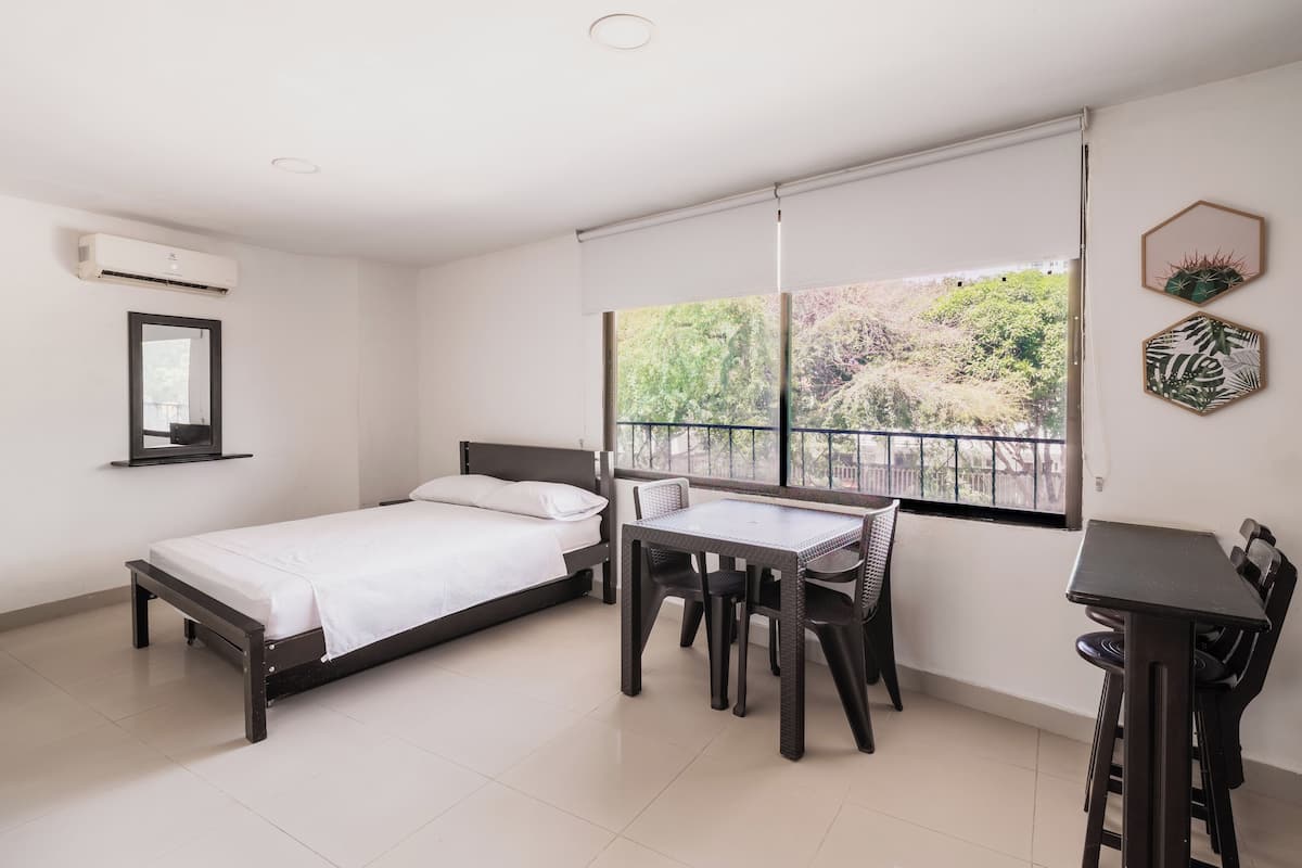 Quarto com cama, escrivaninha e cadeira, janela com vista para árvores e ar condicionado.
