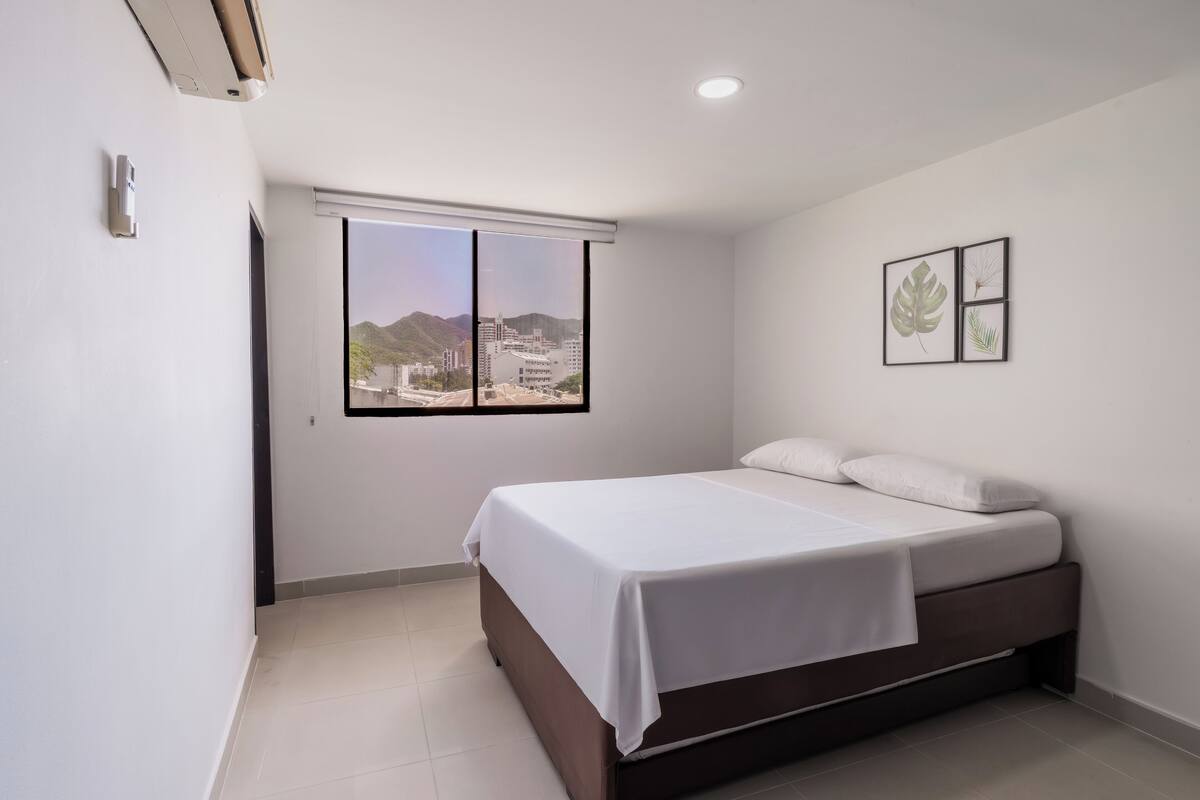 Quarto com cama, janela com vista para edifícios e montanhas, e ar condicionado fixado na parede.
