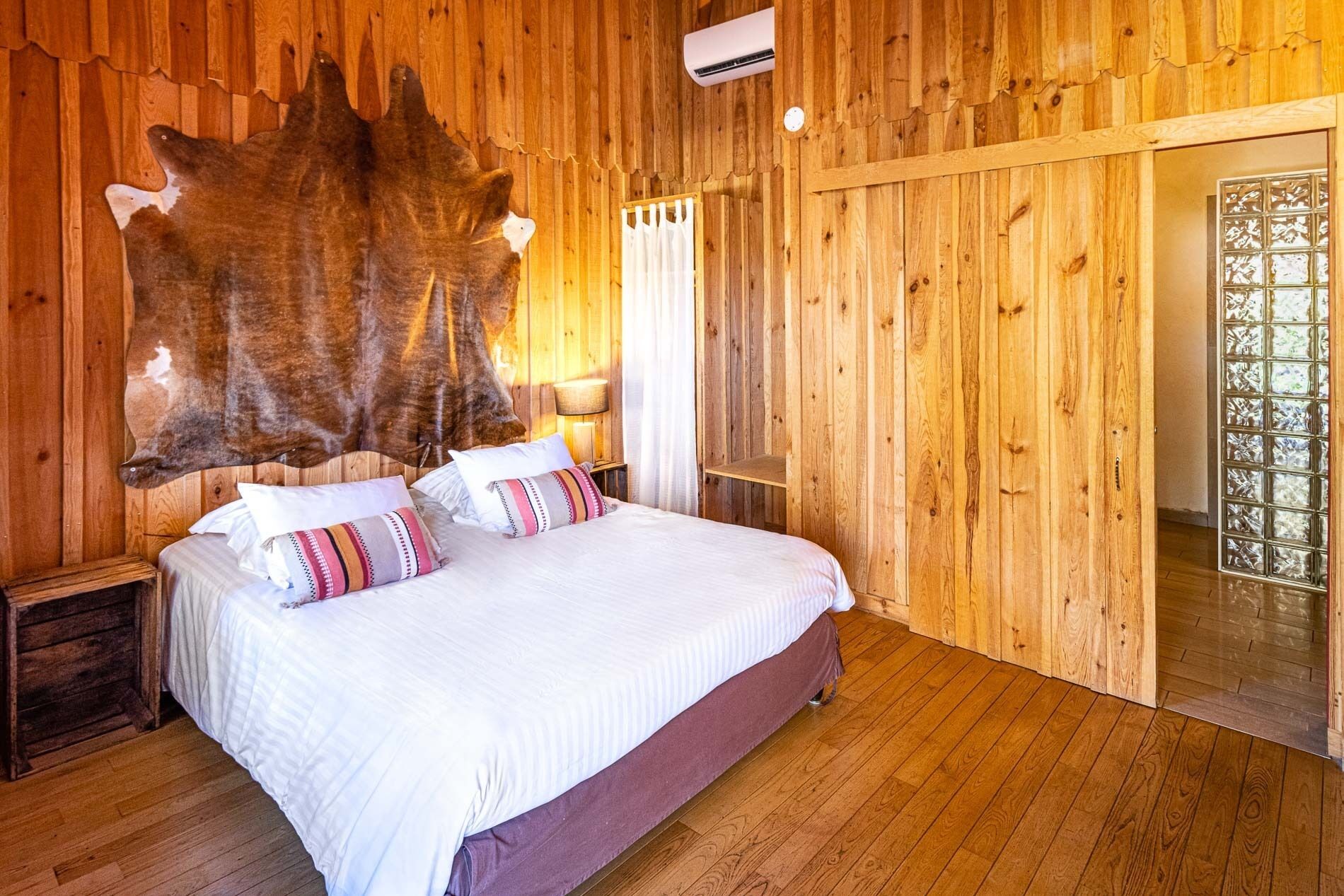 Um quarto de madeira com um tapete de pele grande, uma cama com roupa de cama branca e almofadas estampadas, uma mesa de cabeceira, um abajur e uma janela com cortinas.