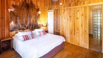 Um quarto de madeira com um tapete de pele grande, uma cama com roupa de cama branca e almofadas estampadas, uma mesa de cabeceira, um abajur e uma janela com cortinas.