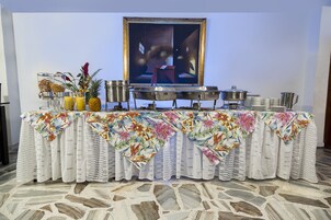 Mesa de buffet com toalha florida, diversos recipientes de comida e um quadro na parede.