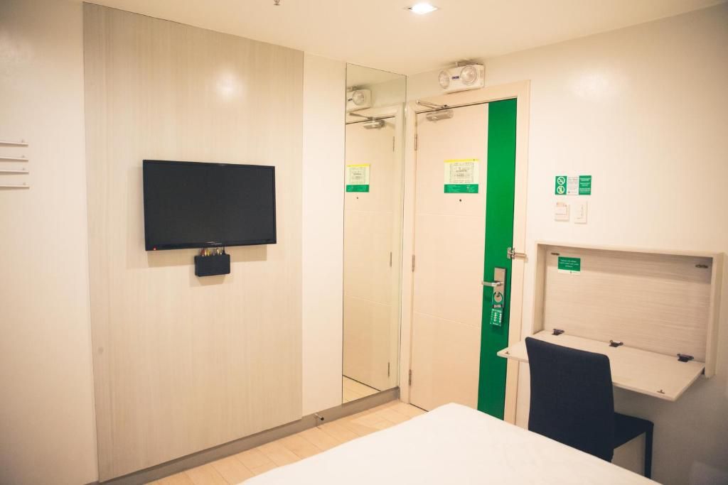 Quarto de hotel com uma cama, televisão na parede, espelho, porta verde e uma pequena escrivaninha com cadeira.