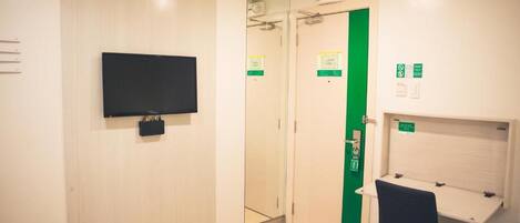 Quarto de hotel com uma cama, televisão na parede, espelho, porta verde e uma pequena escrivaninha com cadeira.
