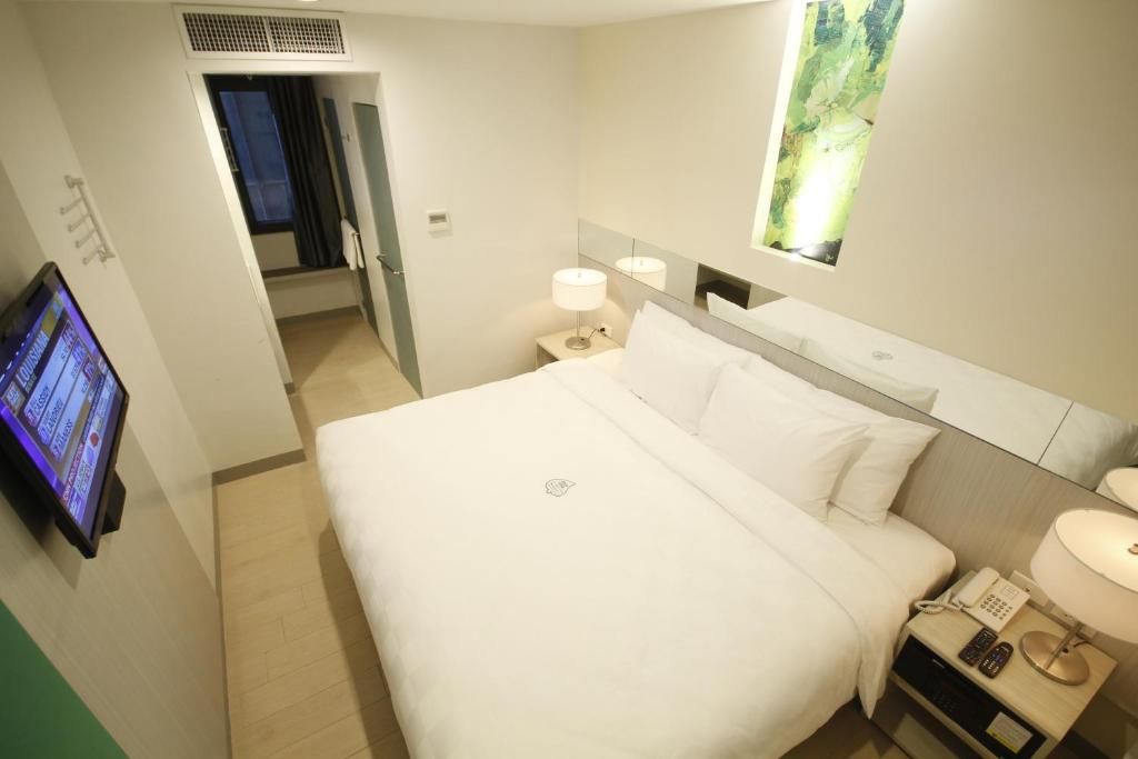 Quarto de hotel com cama, mesa de cabeceira com abajur, telefone, televisão na parede e janela com vista.