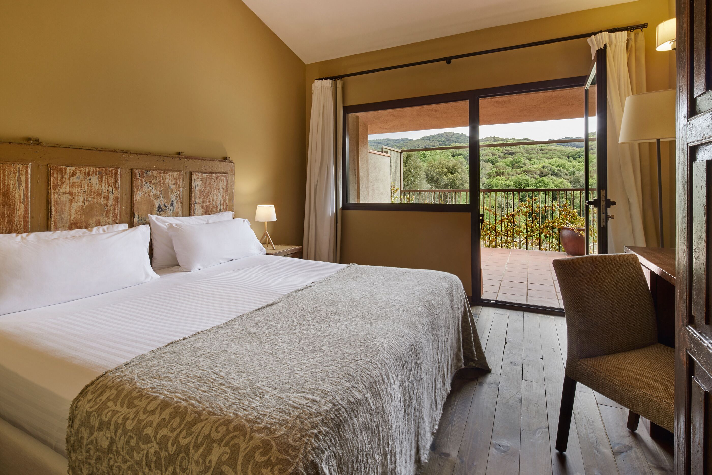 Quarto de hotel com uma cama grande, uma cadeira, varanda com vista para vegetação e janela com cortinas.
