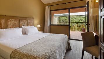 Quarto de hotel com uma cama grande, uma cadeira, varanda com vista para vegetação e janela com cortinas.