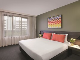 Quarto de hotel com uma cama grande, uma mesa de cabeceira, uma luminária, uma janela com cortinas leves e um quadro abstrato colorido na parede.