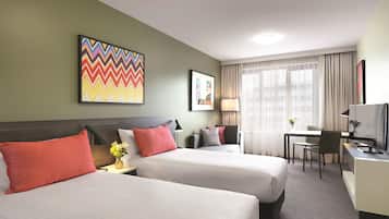 Quarto de hotel com duas camas, uma mesa, uma cadeira, televisão e janela com cortinas.