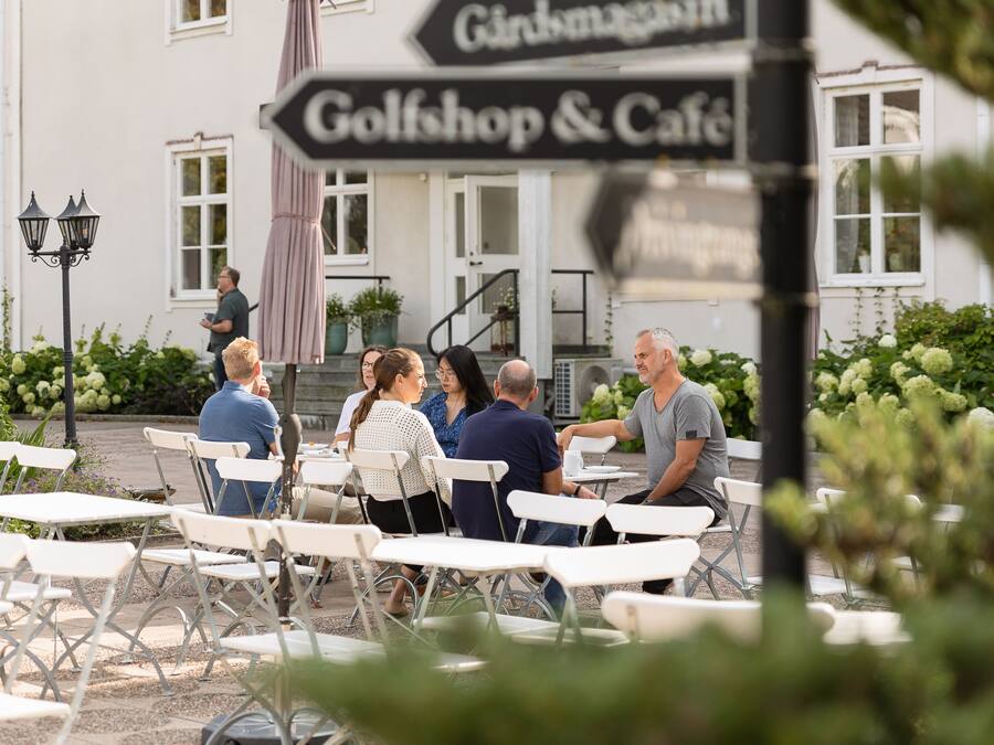 Uma área de estar externa com mesas e cadeiras, um edifício com uma placa indicando "Golfshop & Café" e uma escadaria que leva a uma porta.