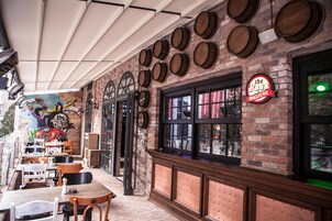 Uma área externa com mesas e cadeiras, paredes de tijolo, barris de madeira e uma placa com a inscrição 'The Rosa Brasserie'.