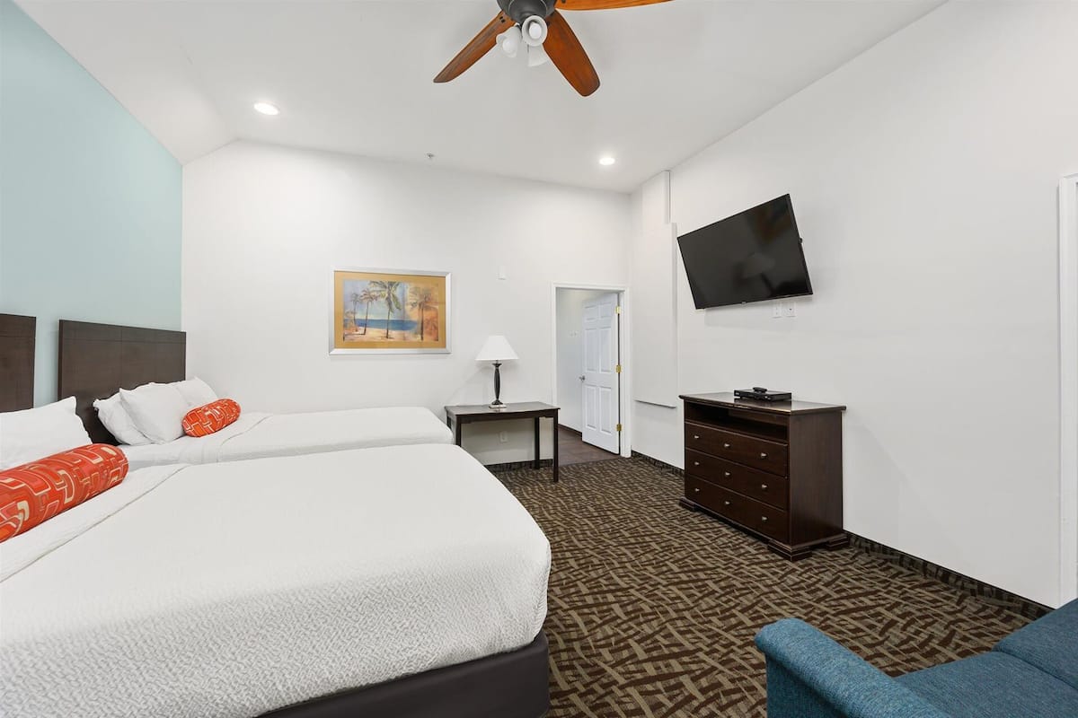 Quarto de hotel com duas camas, um ventilador de teto, televisão, escrivaninha e tapete estampado.