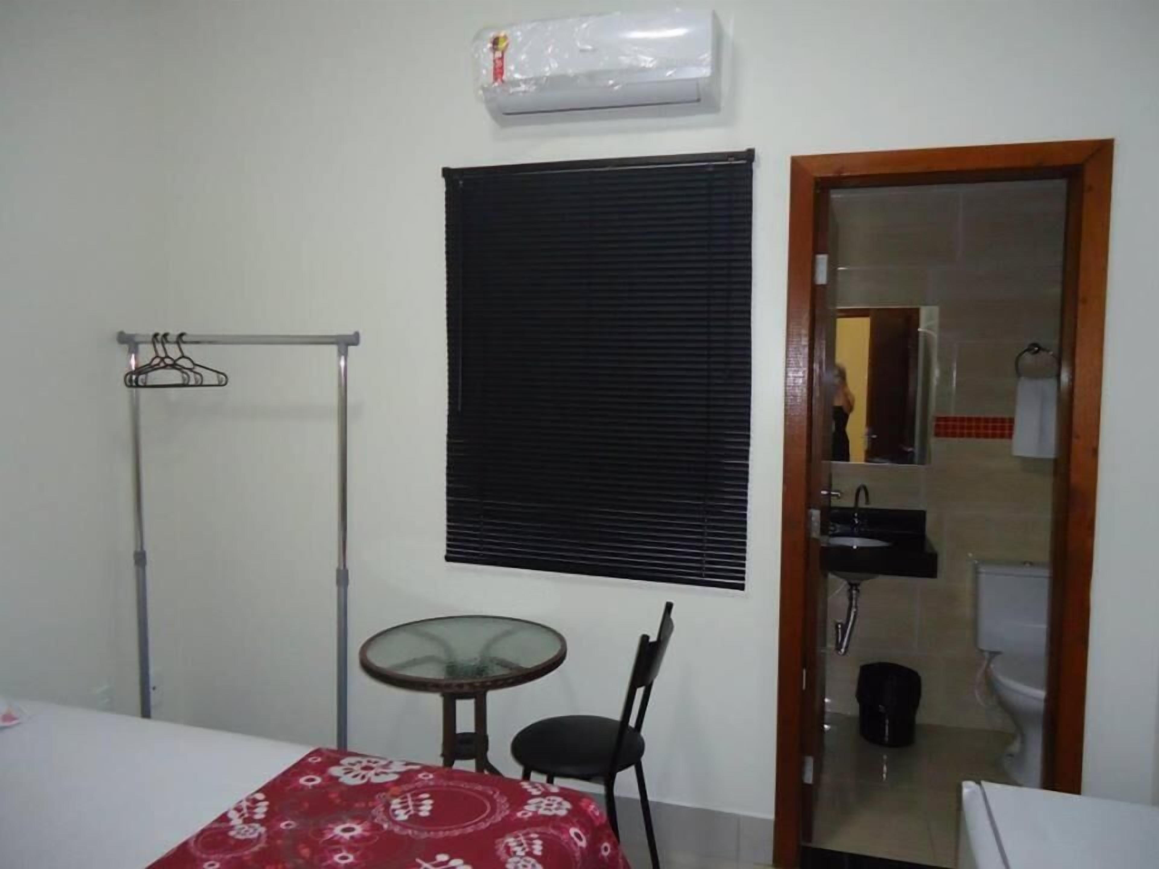 Quarto de hotel com cama, uma pequena mesa com cadeira, janela com persianas, cabideiro e banheiro visível por uma porta aberta.