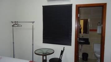 Quarto de hotel com cama, uma pequena mesa com cadeira, janela com persianas, cabideiro e banheiro visível por uma porta aberta.