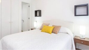 Quarto de hotel com cama, mesa de cabeceira, prateleira na parede e uma porta.