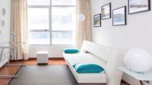 Quarto moderno com sofá branco, almofadas verde-água, um banquinho branco pequeno, luminária branca e quadros nas paredes.