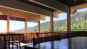 Restaurante com mesas e cadeiras de madeira, grandes janelas com vista para uma montanha arborizada.