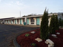 Um motel com fileira de quartos, estacionamento e área ajardinada com plantas e pedras.