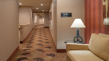Um corredor de hotel com carpete estampado, um sofá, uma luminária e placas indicativas do elevador.
