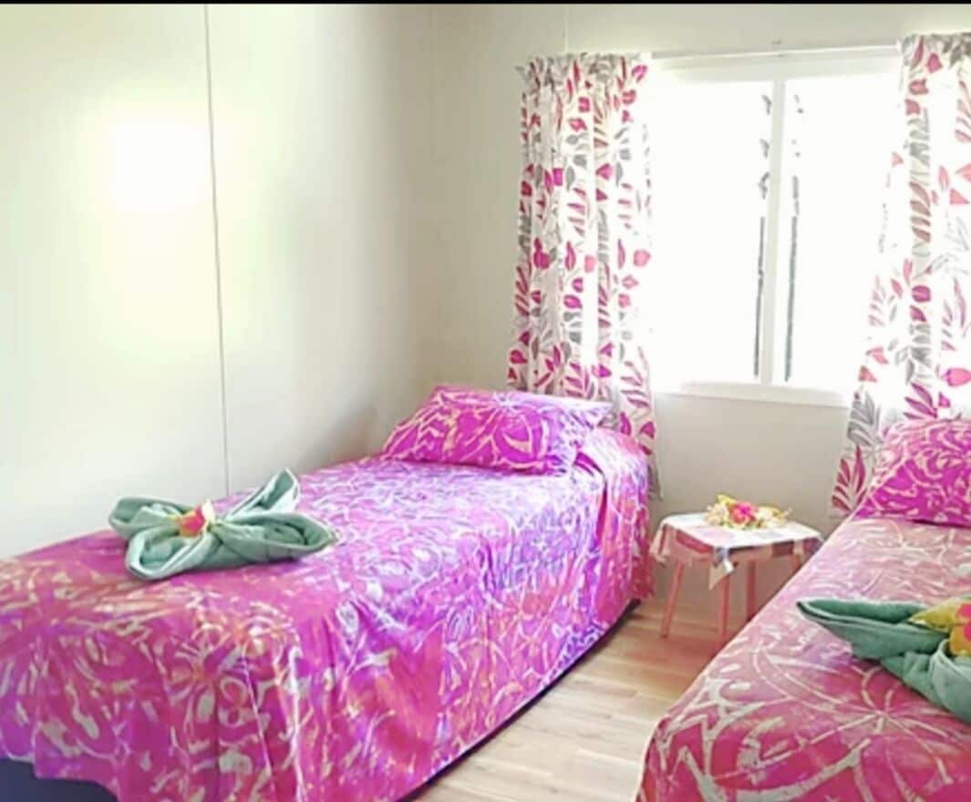 Quarto com duas camas, cada uma com colcha e almofadas estampadas com flores rosa, uma pequena mesa de madeira com arranjo de flores e uma janela com cortinas padronizadas.