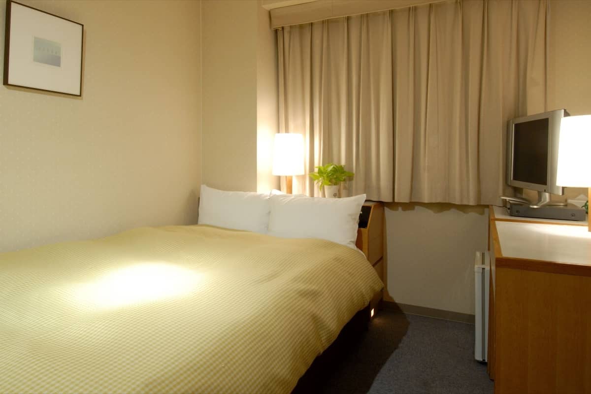 Quarto de hotel com cama, escrivaninha, televisão, luminária e pequeno frigorífico.