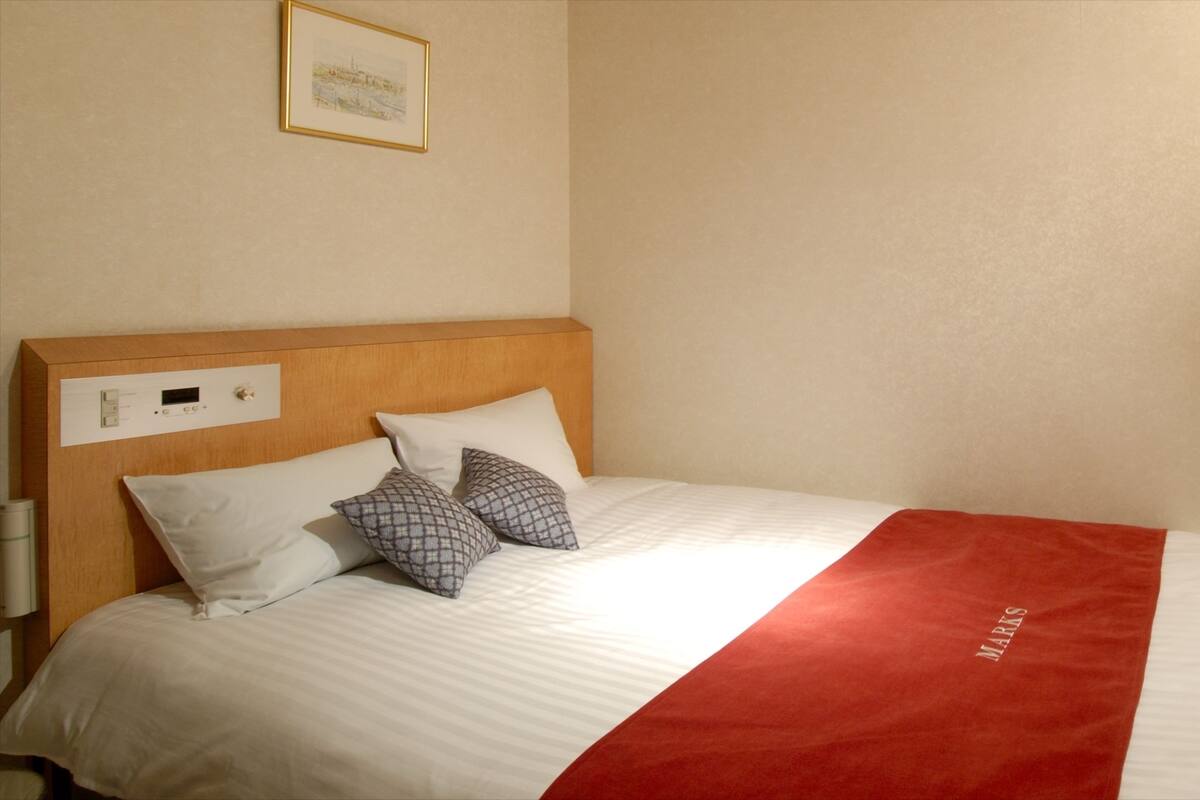 Cama bem arrumada com um cobertor vermelho, uma televisão sobre uma mesa de madeira e um quadro pendurado na parede.