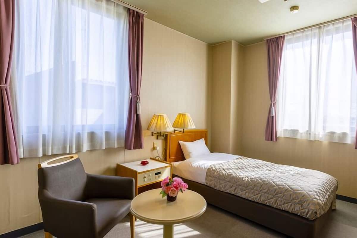 Quarto de hotel com cama, uma cadeira, uma mesinha com um vaso de flores e duas luminárias em uma mesinha de cabeceira.