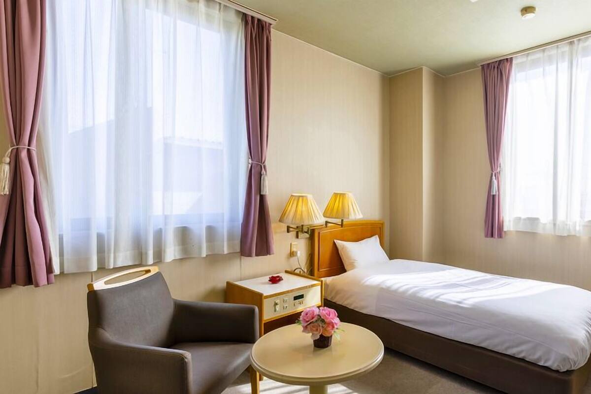 Quarto de hotel com cama, uma cadeira, uma mesinha com um vaso de flores e duas luminárias em uma mesa de cabeceira.