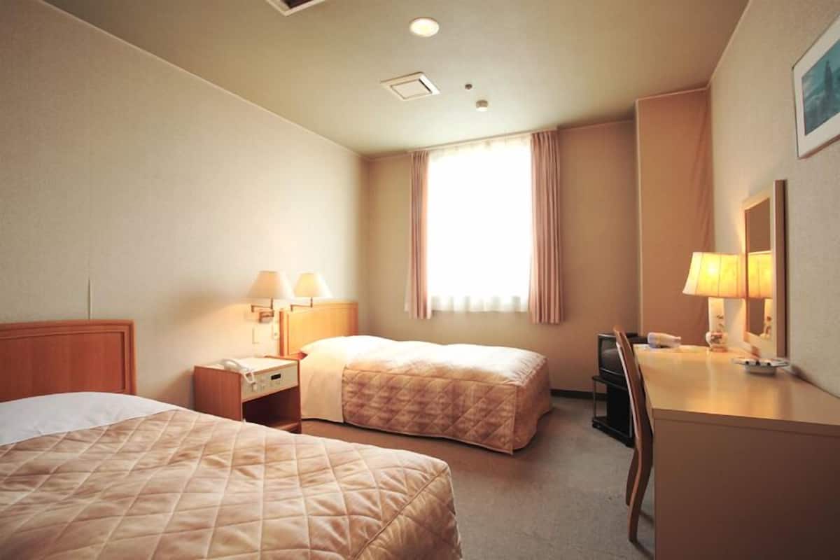 Quarto de hotel com duas camas, uma escrivaninha com cadeira, telefone e janela com cortinas.