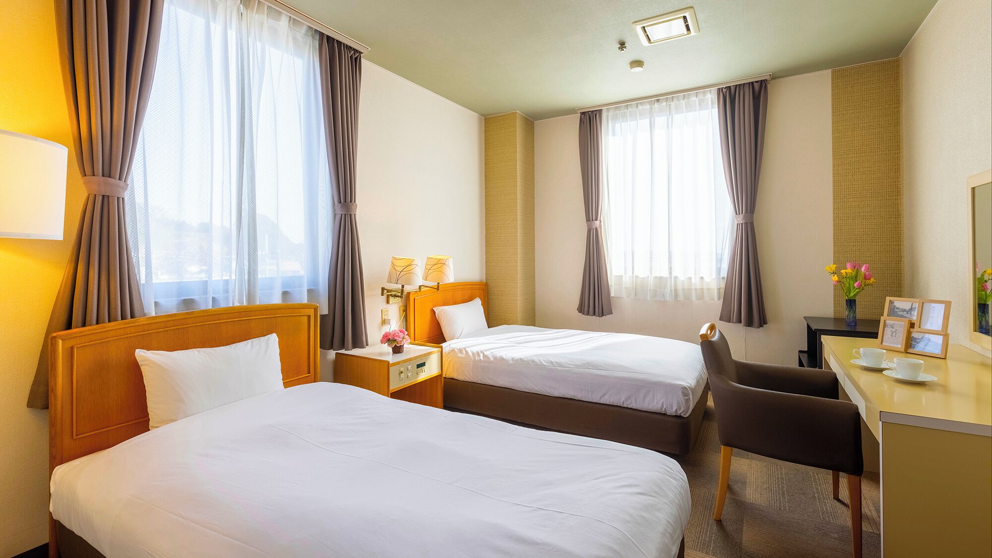 Quarto de hotel com duas camas, uma escrivaninha, uma cadeira e uma pequena mesa com um vaso de flores.