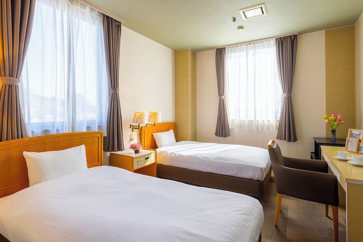 Quarto de hotel com duas camas, uma escrivaninha, uma cadeira e uma pequena mesa com um vaso de flores.