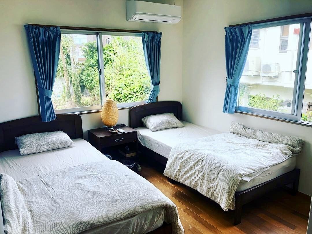 Quarto com duas camas de solteiro, uma mesinha com abajur e uma janela com cortinas.