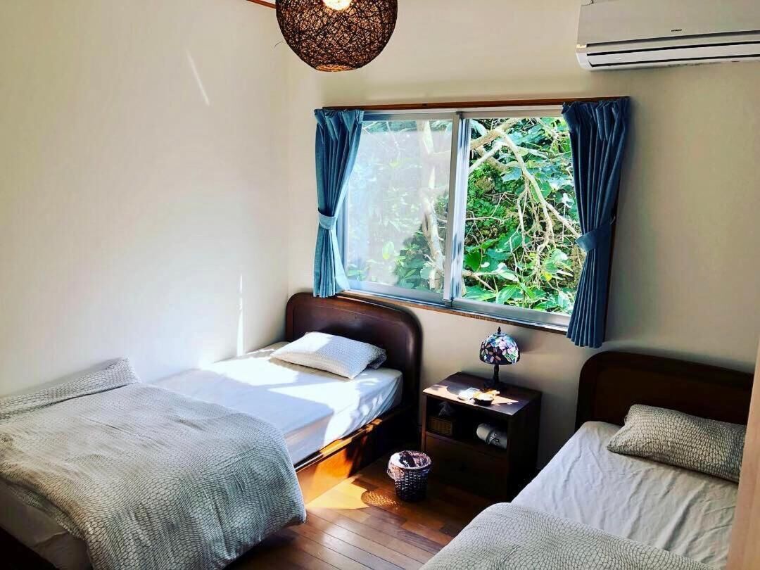 Quarto com duas camas, janela com cortinas, mesa de cabeceira com abajur e ar condicionado.