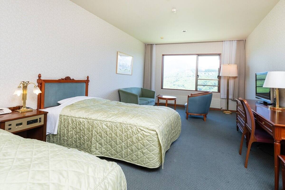 Quarto de hotel com duas camas, uma escrivaninha, uma cadeira, uma televisão e uma janela com vista.