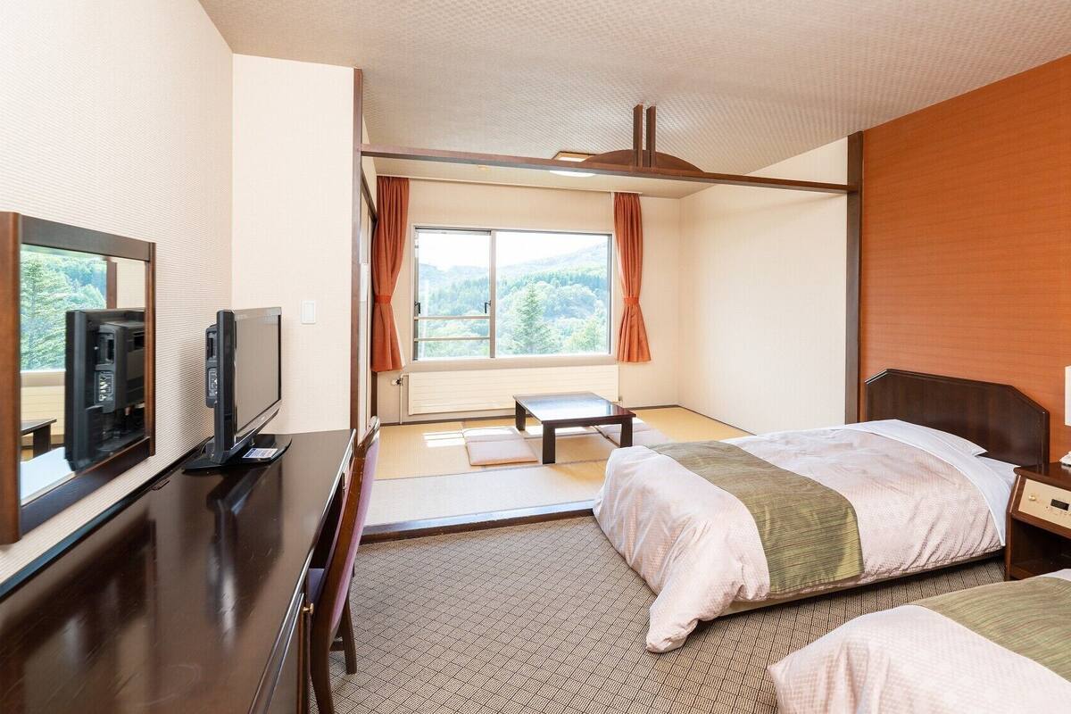 Quarto de hotel com duas camas, uma mesa com televisão, uma janela com cortinas e uma mesa de cabeceira com abajur, além de vista para o exterior.