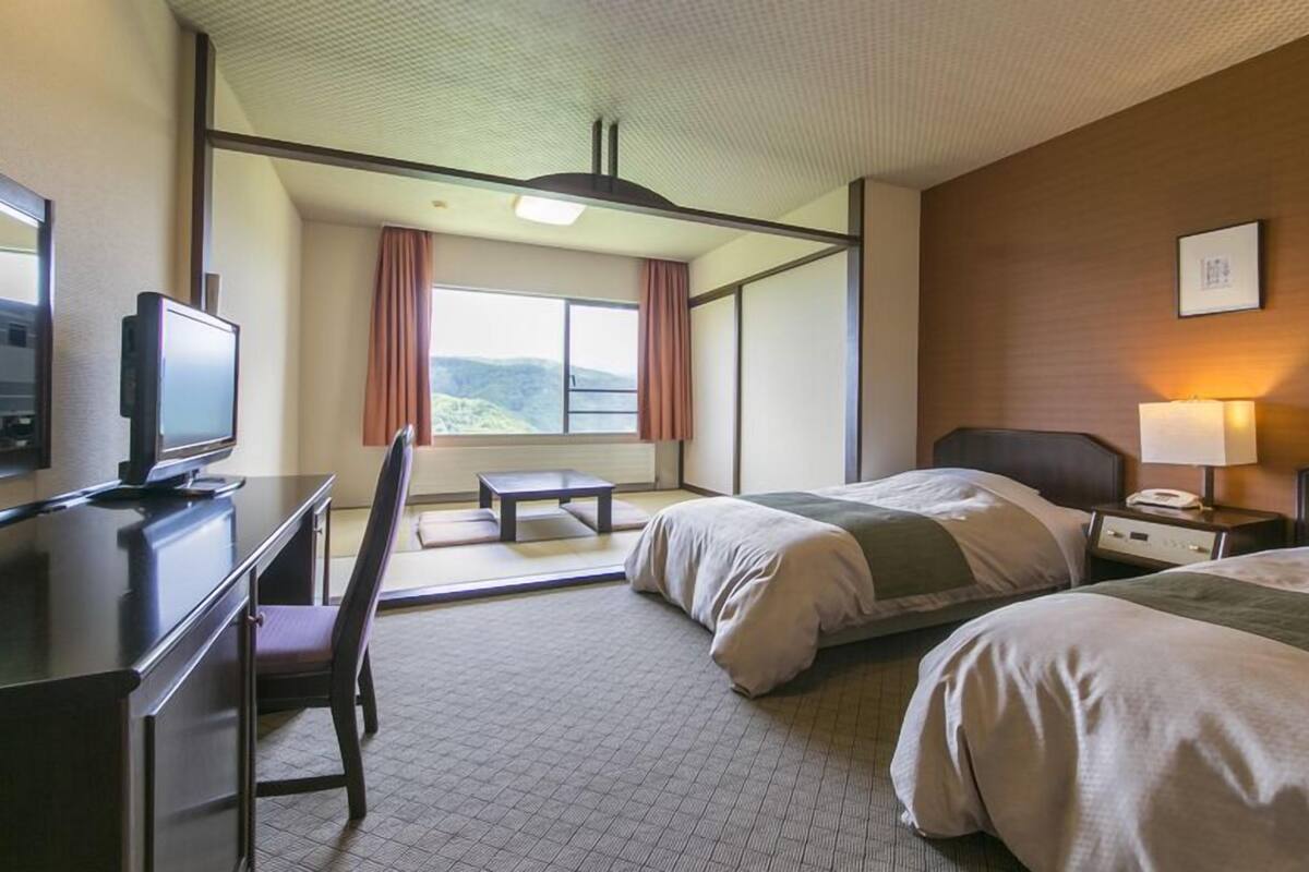 Quarto de hotel com duas camas, uma escrivaninha com cadeira, televisão, janela com cortinas e mesa de cabeceira com abajur.