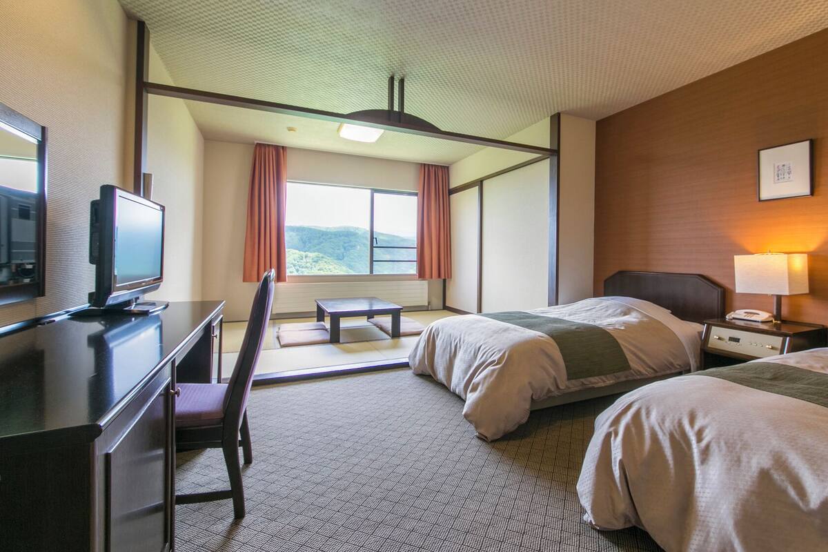 Quarto de hotel com escrivaninha, duas camas, televisão, janela com cortinas e luminária.