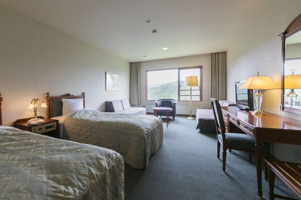 Quarto de hotel com duas camas, uma escrivaninha, uma cadeira, um abajur e uma janela grande.