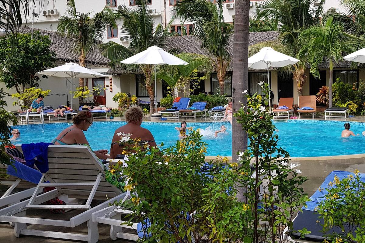 Área de piscina com várias espreguiçadeiras e guarda-sóis, cercada por palmeiras e um edifício com telhado telhado.