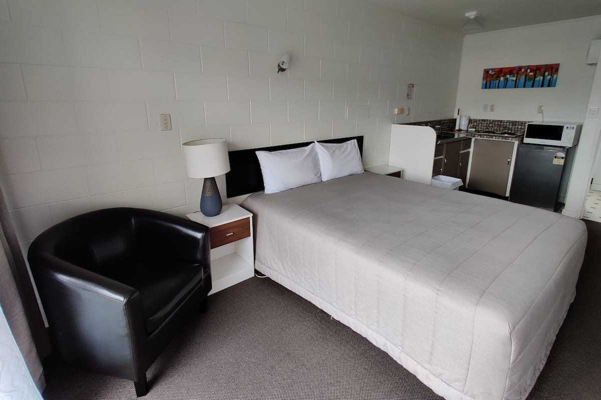 Quarto de hotel com cama, poltrona, mesa de cabeceira, luminária, micro-ondas e geladeira.