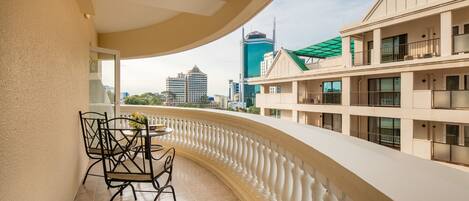 Uma varanda com balaústres brancos curvos, uma mesa com cadeiras e vista para um horizonte urbano com edifícios modernos.