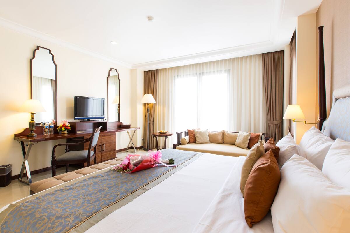 Quarto de hotel com uma cama grande, uma escrivaninha com televisão, uma cadeira e um sofá.
