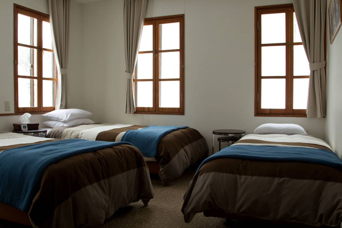 Quarto de hotel com três camas, janelas amplas e uma pequena mesa.