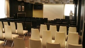 Uma sala de conferência com fileiras de cadeiras, uma tela de projeção e uma televisão na parede.