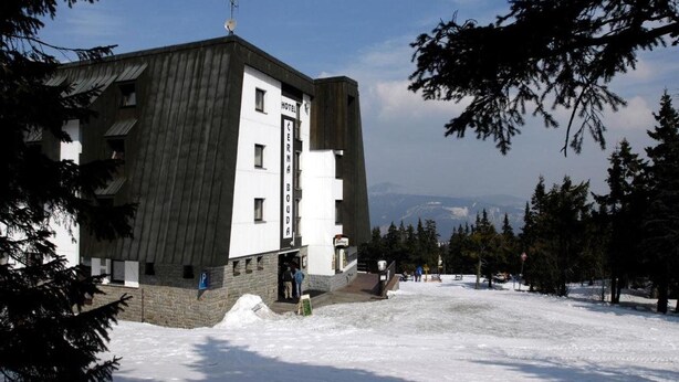 Hotel com fachada branca e telhado escuro, cercado por neve e árvores, com vista para montanhas ao fundo.