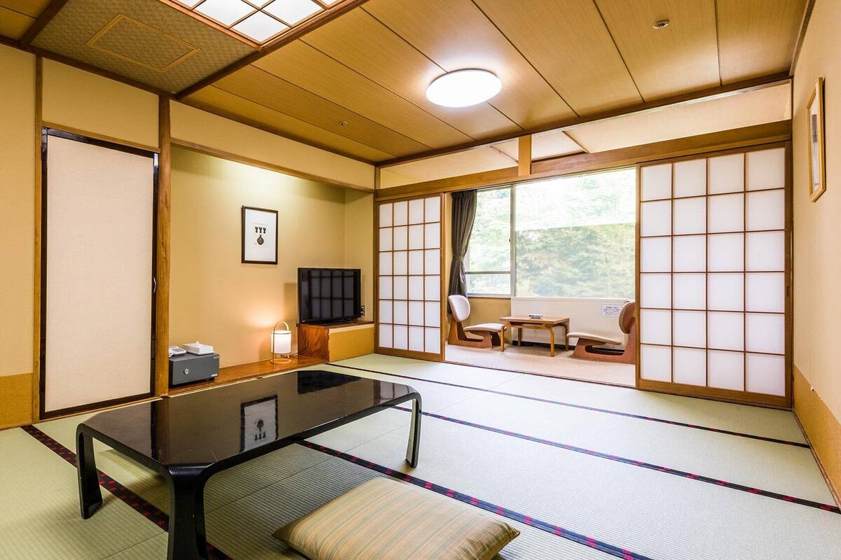 Um quarto japonês tradicional com tatames, uma mesa baixa, uma televisão e portas deslizantes que dão para uma varanda.