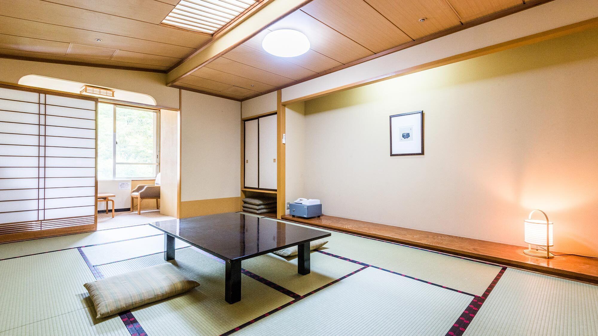 Um espaço com tatames, uma mesa baixa, um assento, um quadro na parede e uma janela com vista.