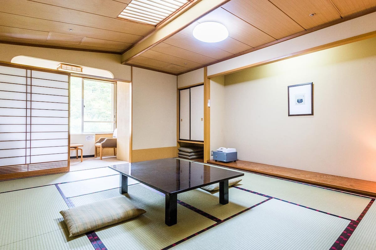 Um espaço com tatames, uma mesa baixa, um assento, um quadro na parede e uma janela com vista.