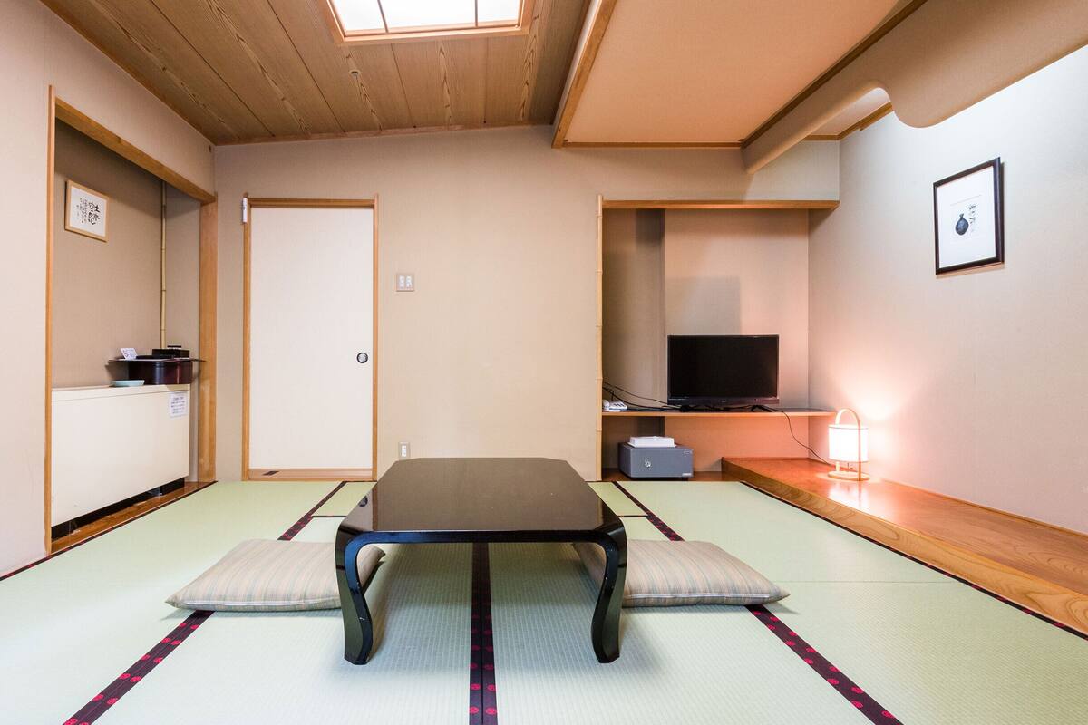 Um quarto tradicional com esteiras de tatame, uma mesa baixa, almofadas, uma televisão e uma pequena escrivaninha com uma luminária.