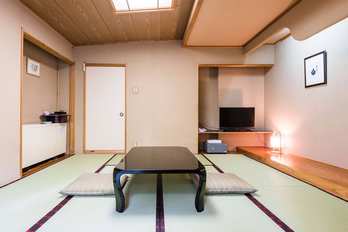 Um quarto tradicional com tatames, uma mesa baixa, almofadas, uma televisão e um pequeno estante com objetos.