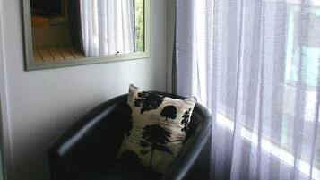 Quarto com uma cadeira de couro preto, um travesseiro branco com um desenho de árvore, um espelho na parede e cortinas leves que permitem a entrada de luz natural.